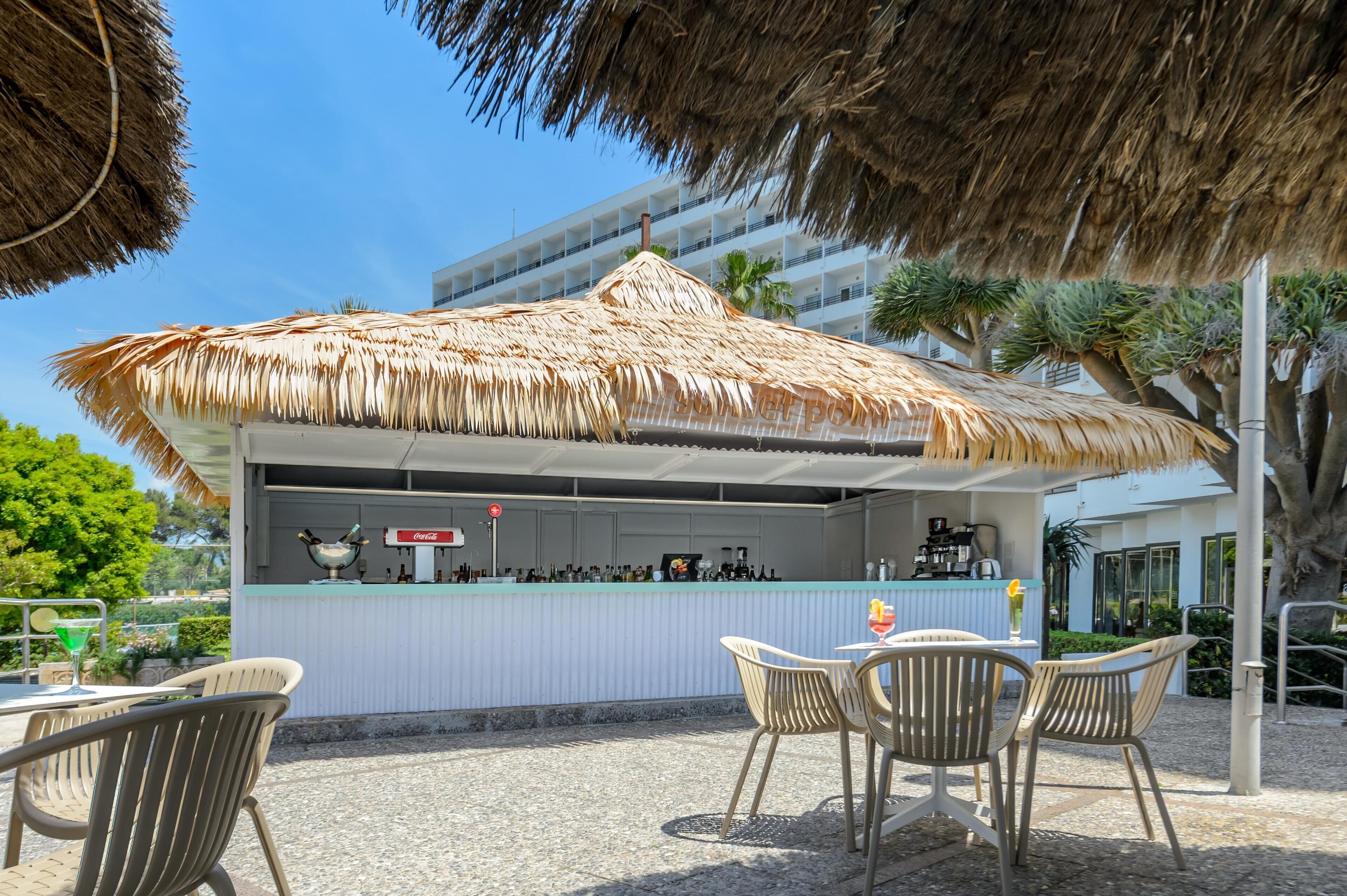 Um bar à beira-mar com telhado de palha, mesas ao ar livre e vista para um edifício alto ao fundo.