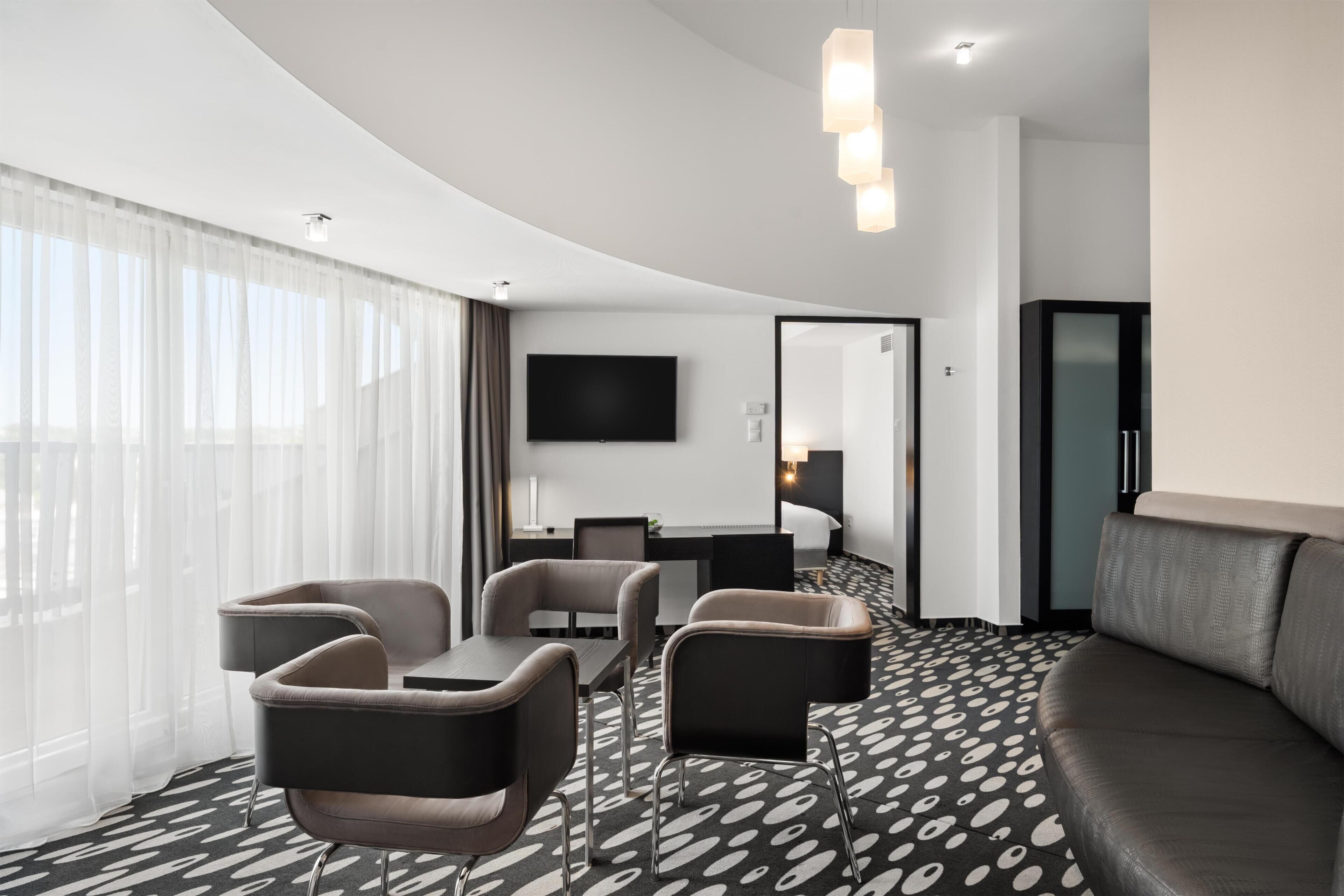 Quarto de hotel moderno com teto curvo, televisão de tela plana, sofá e área de jantar com cadeiras e mesa.