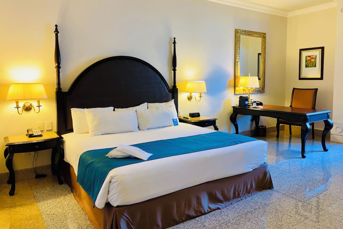 Quarto de hotel com cama de dossel, duas luminárias de cabeceira, uma escrivaninha, uma cadeira e um espelho grande na parede.