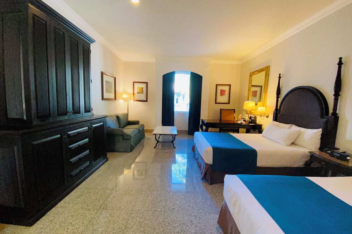 Quarto de hotel com duas camas, uma escrivaninha, um sofá, um guarda-roupa e um espelho.