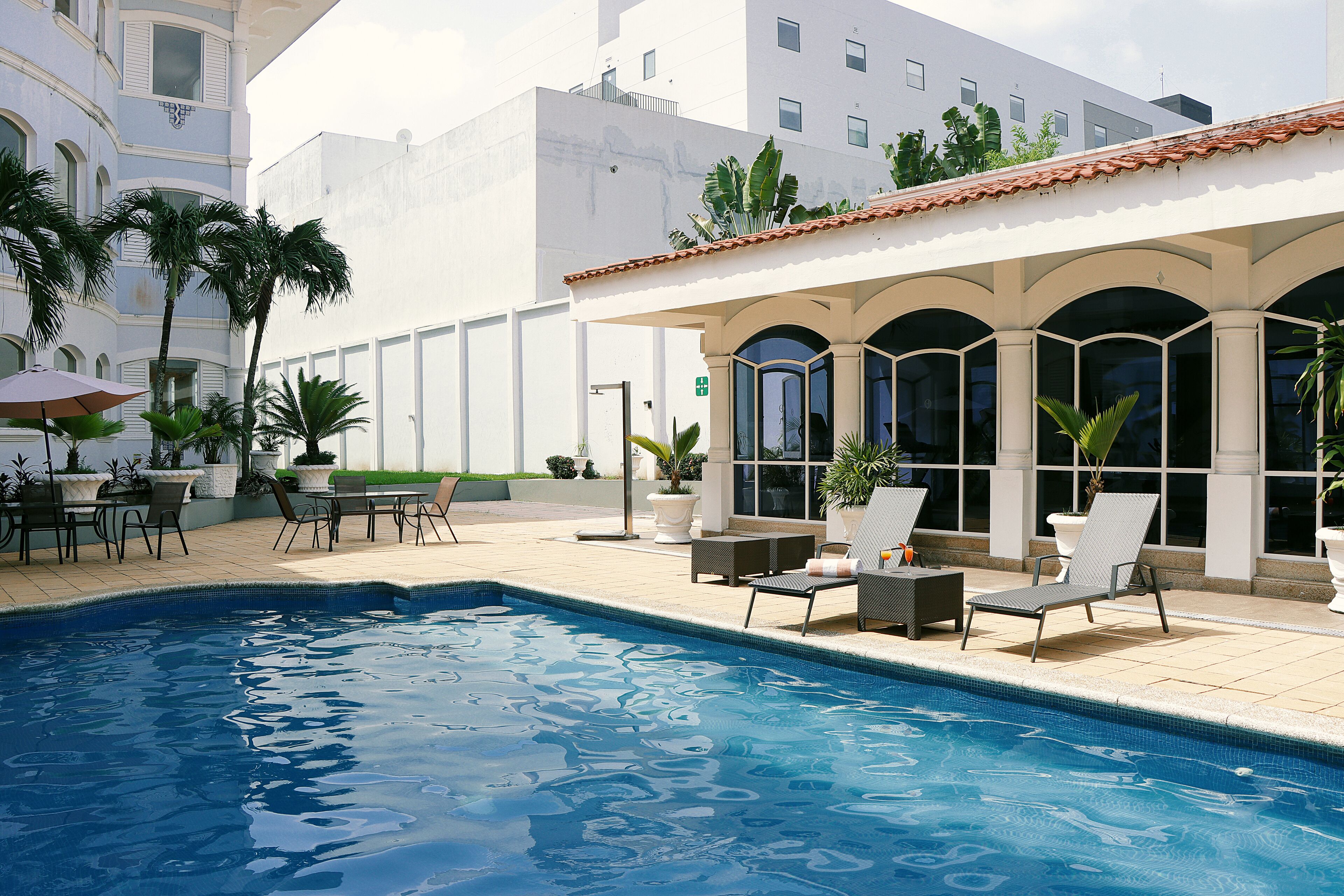 Piscina com espreguiçadeiras e mesas, cercada por plantas em vasos e um edifício com janelas em arco.