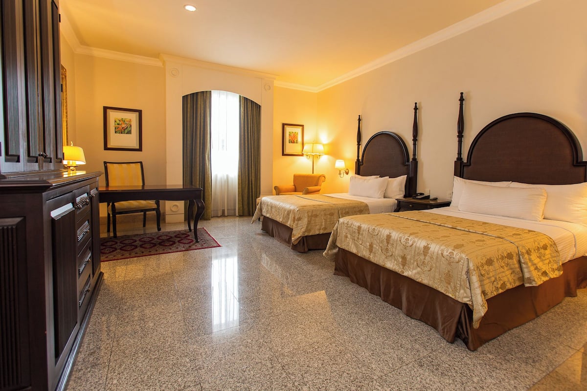 Quarto de hotel com duas camas, uma escrivaninha, uma cadeira e um aparador.