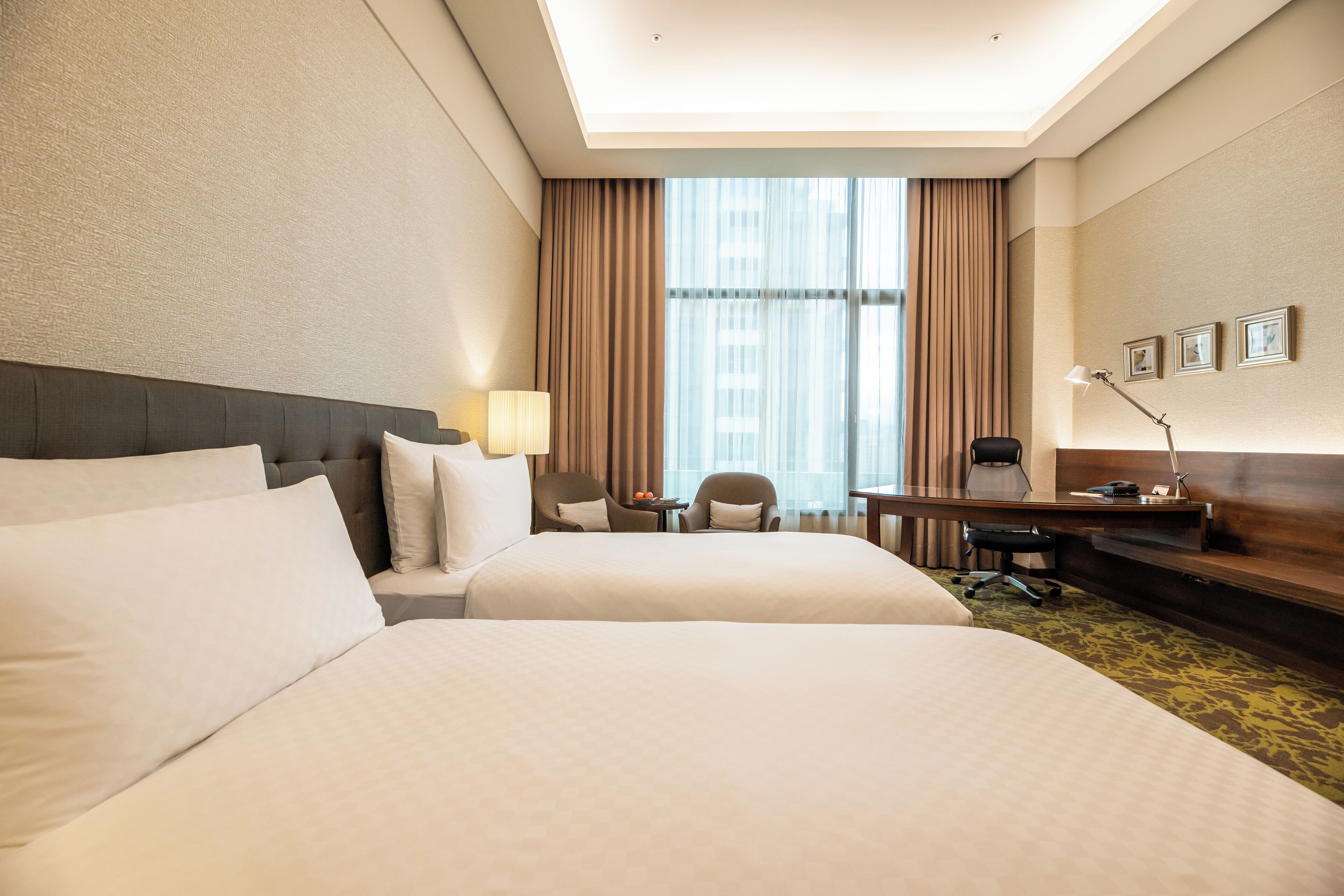 Quarto de hotel com duas camas, uma escrivaninha, uma cadeira, uma luminária e vista da cidade por grandes janelas.