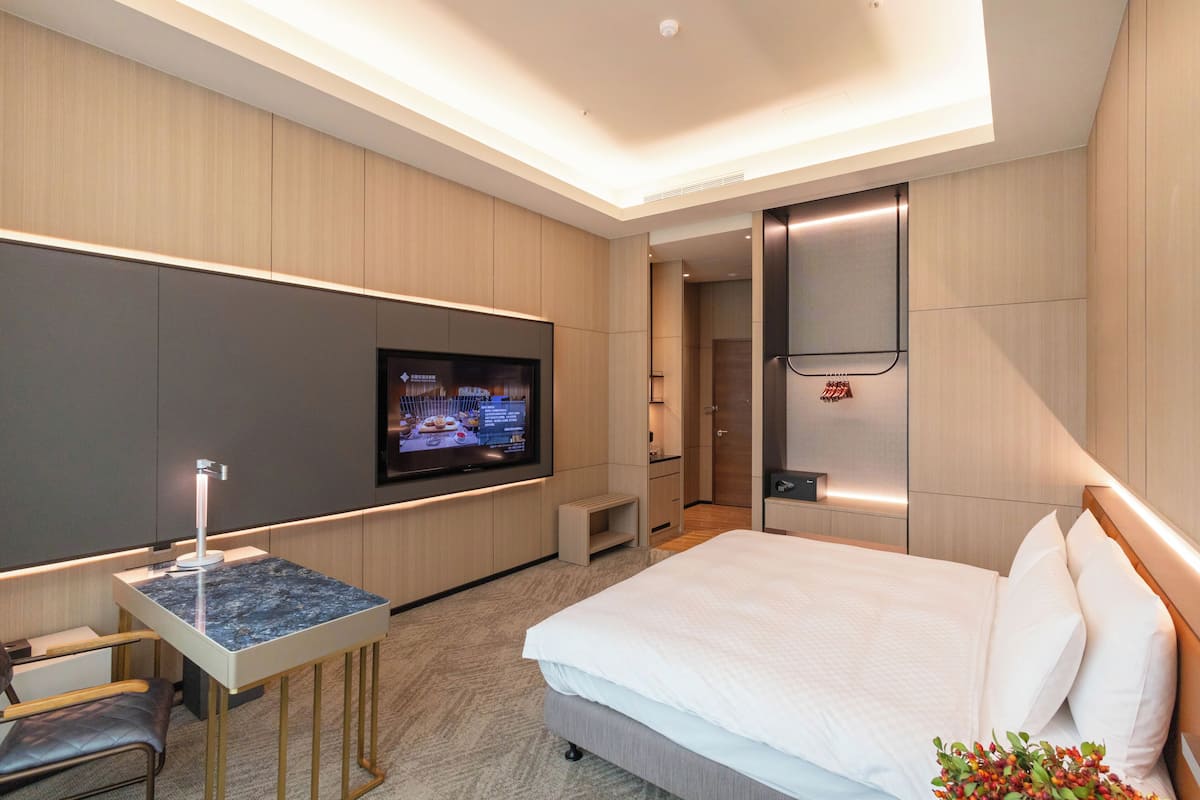 Quarto de hotel moderno com uma cama grande, televisão de tela plana na parede, uma pequena escrivaninha com luminária e um armário embutido.