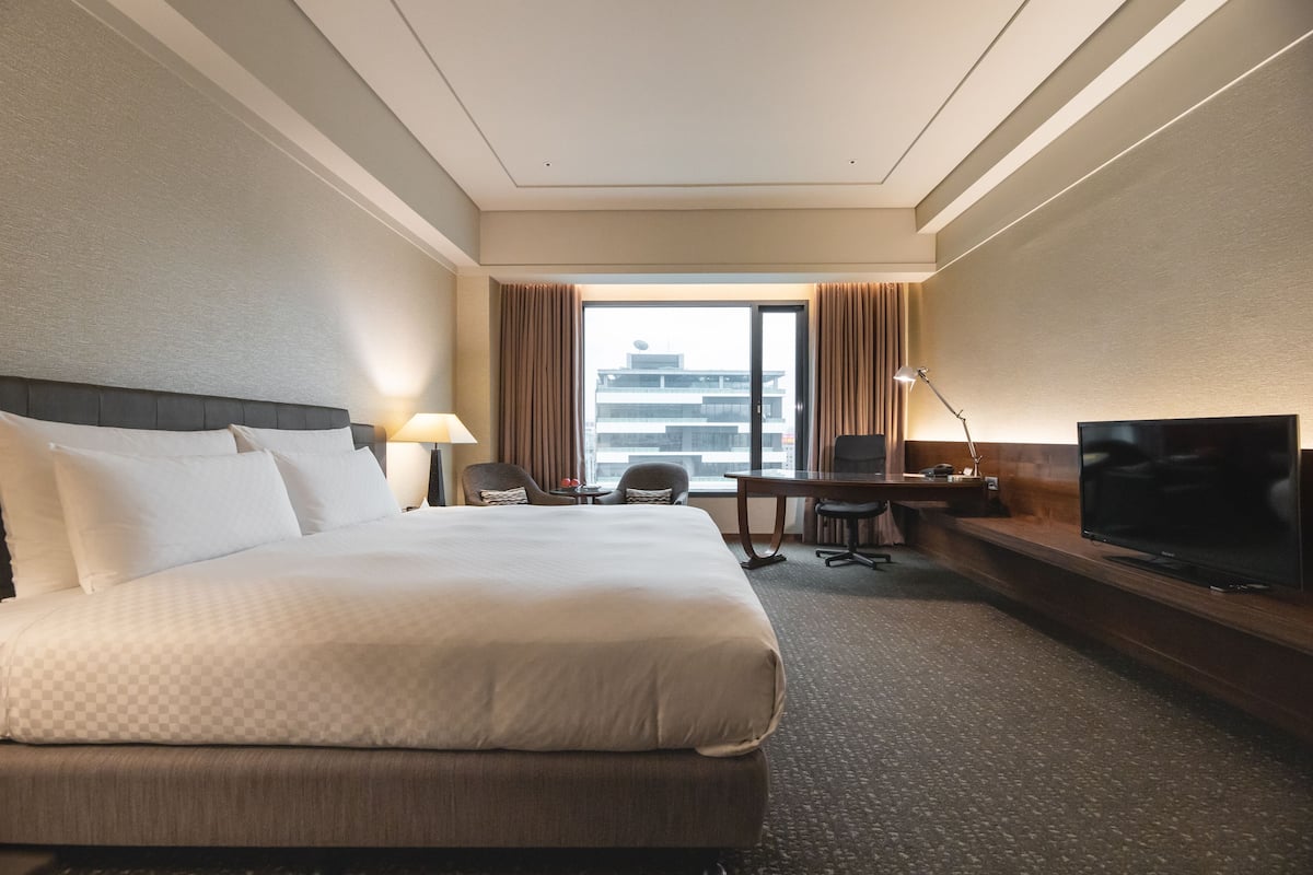 Quarto de hotel com uma cama grande, uma escrivaninha com computador, televisão e vista para um edifício pela janela.