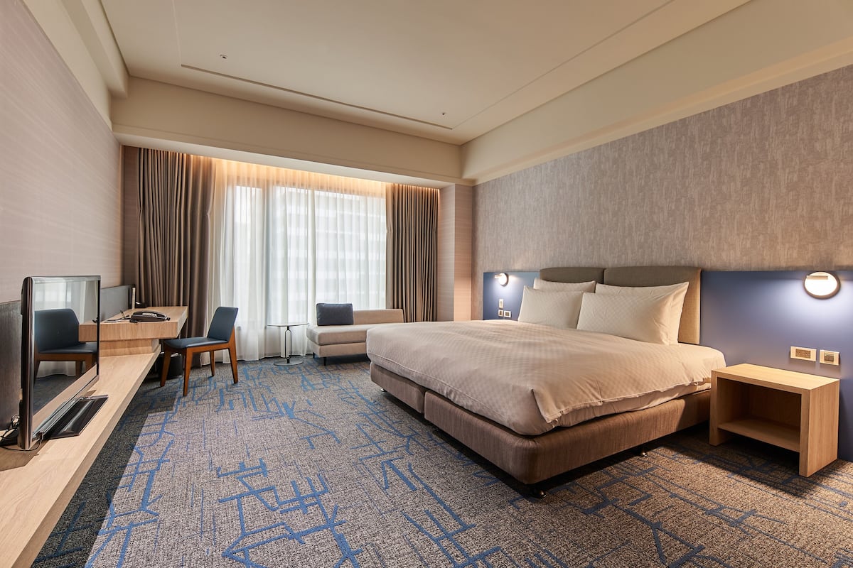 Quarto de hotel com uma cama grande, uma escrivaninha com cadeira, televisão e uma mesinha.