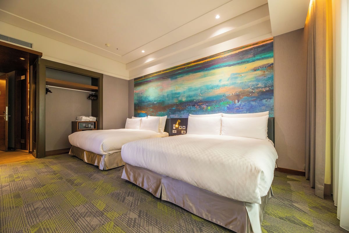 Quarto de hotel com duas camas, um quadro abstrato grande na parede e piso coberto por carpete.