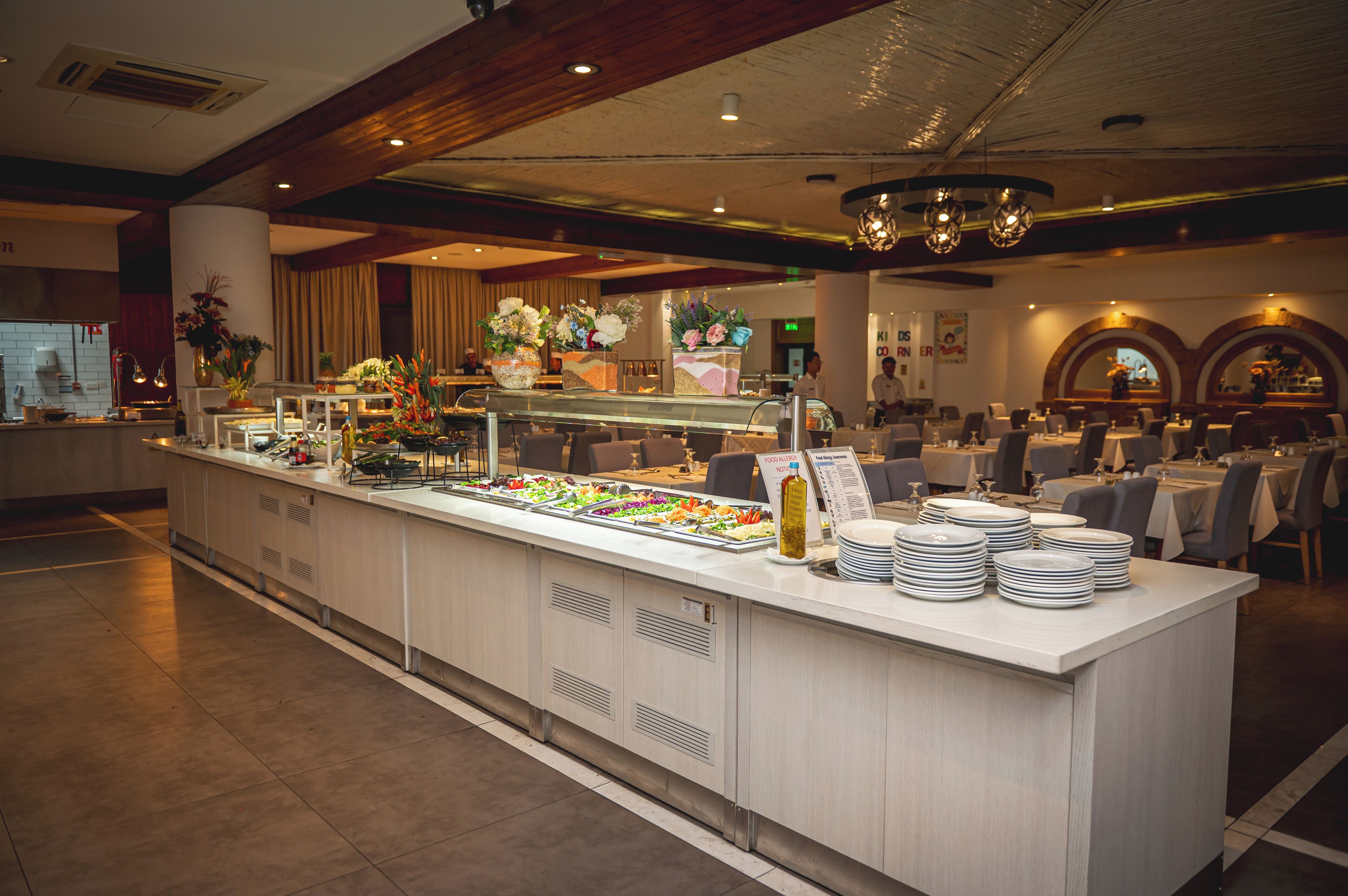 Buffet com diversos pratos, balcão com pratos e área de refeições com mesas e cadeiras.