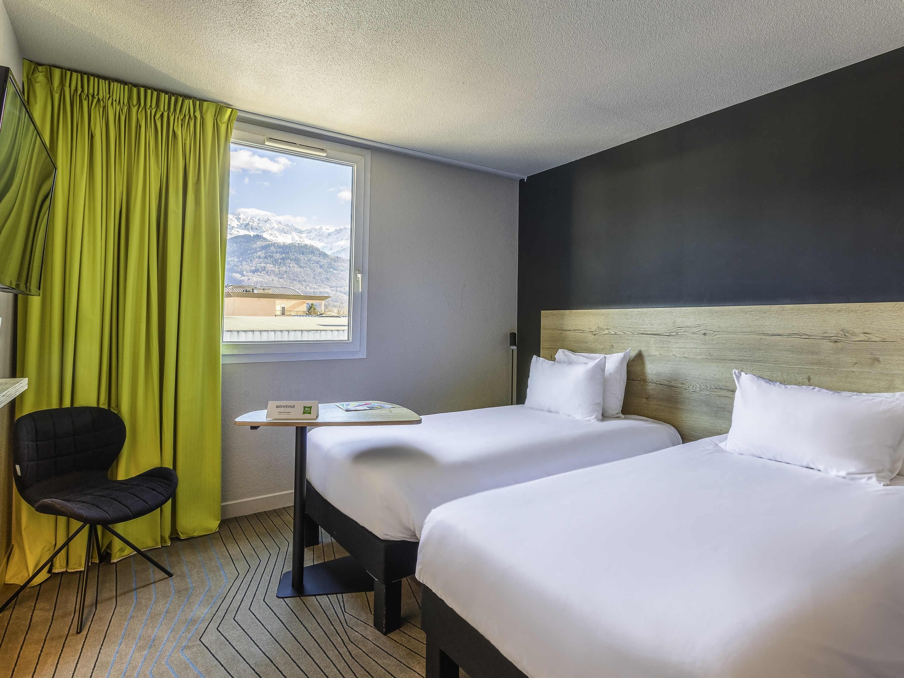 Quarto de hotel com duas camas, uma mesa, uma cadeira e janela com vista para montanhas.