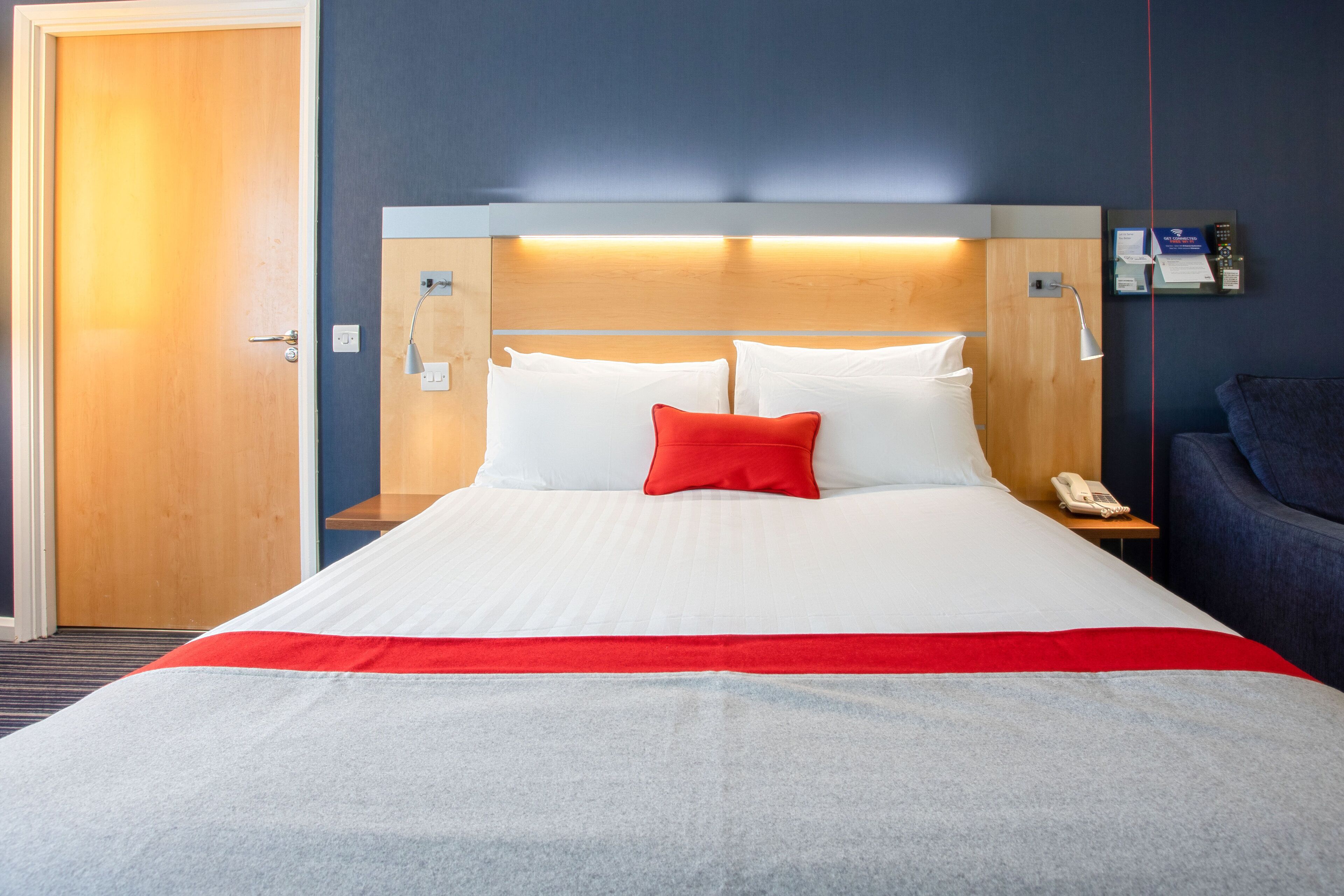 Quarto de hotel com uma cama grande, cabeceira de madeira, travesseiro decorativo vermelho, mesa de cabeceira com abajur e telefone.