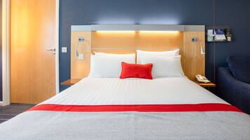 Quarto de hotel com uma cama grande, cabeceira de madeira, travesseiro decorativo vermelho, mesa de cabeceira com abajur e telefone.