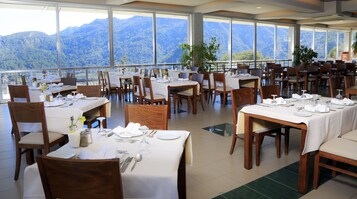 Um restaurante com várias mesas preparadas para refeições, amplas janelas com vista para as montanhas e cadeiras de madeira.
