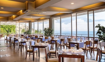 Restaurante com mesas preparadas para refeições, amplas janelas com vista para o mar e plantas em vasos.