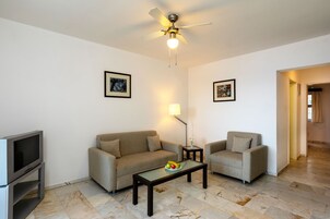 Uma sala de estar com sofá, duas poltronas, uma mesa de centro com frutas, televisão, ventilador de teto e quadros nas paredes.