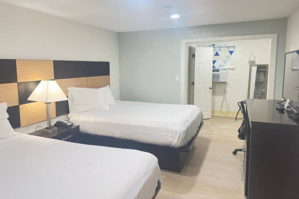 Quarto de hotel com duas camas, uma escrivaninha, uma luminária e um guarda-roupa.