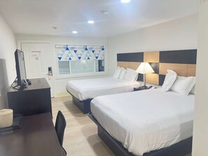 Quarto de hotel com duas camas, uma escrivaninha, uma cadeira, um abajur e uma janela com cortinas geométricas.