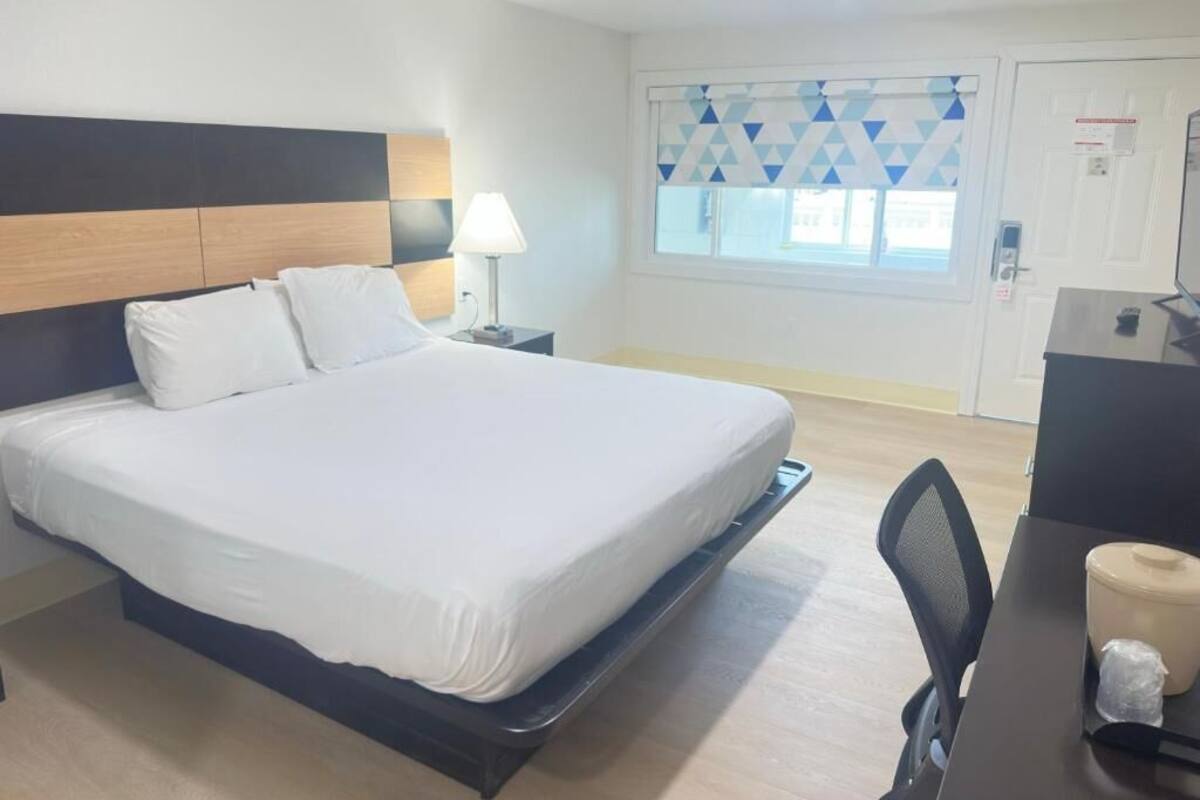 Quarto de hotel com cama, mesa de cabeceira, luminária, janela com persianas e escrivaninha com cadeira.