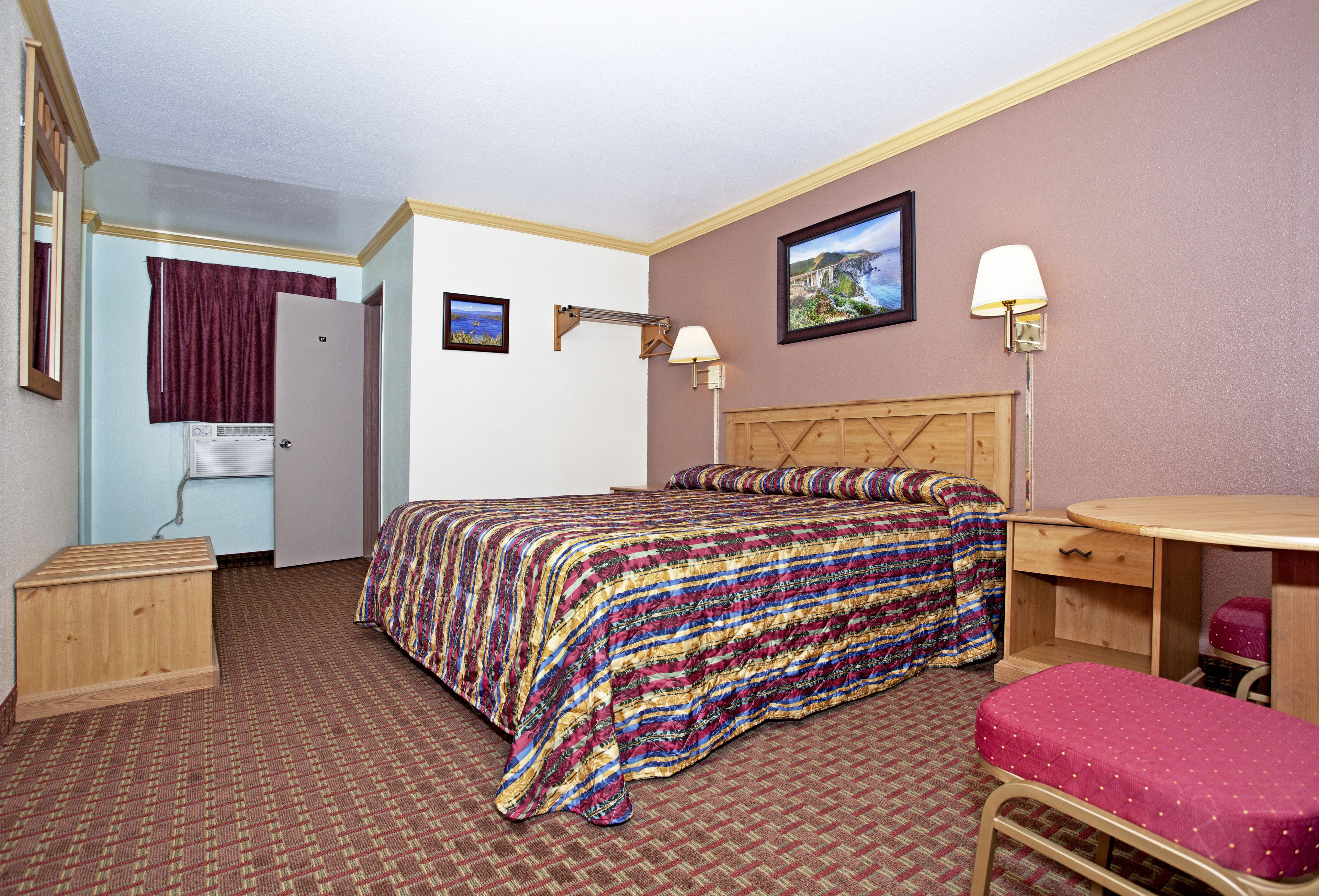 Quarto de hotel com cama, mesa de cabeceira, escrivaninha, cadeira e um pequeno armário de madeira.