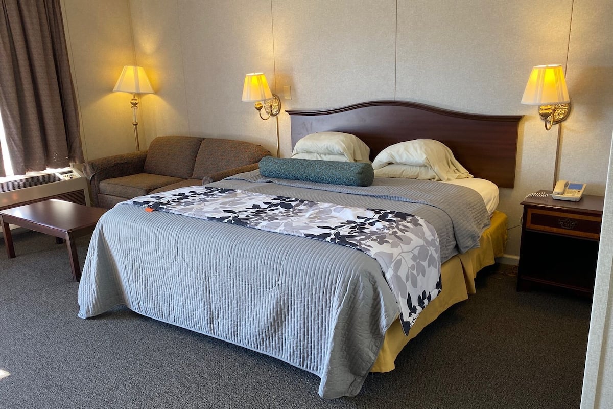 Quarto de hotel com uma cama, duas luminárias laterais, mesa de cabeceira com telefone, uma cadeira e uma escrivaninha.