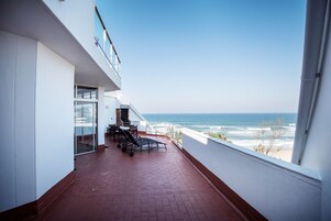Uma varanda com vista para o oceano, uma porta de vidro que dá para o interior e uma mesa pequena com cadeiras.