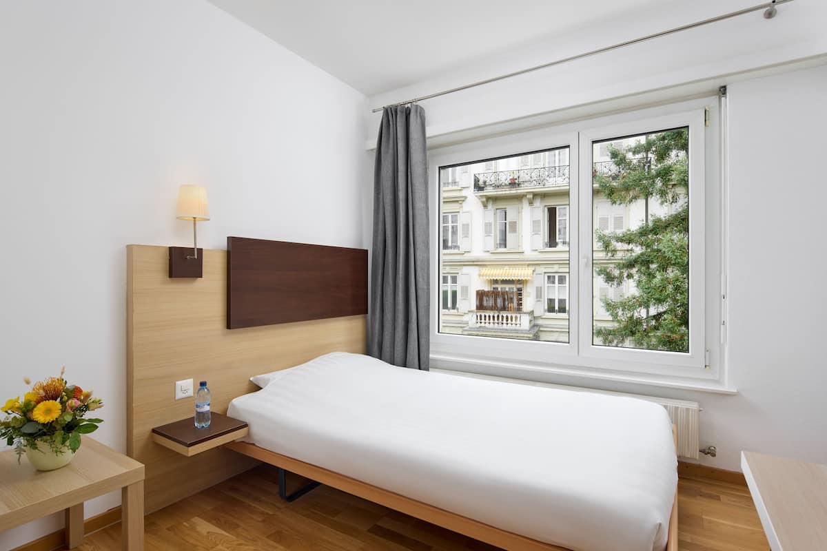 Quarto de hotel com cama, mesa de cabeceira, luminária e janela com vista para edifícios e árvores.