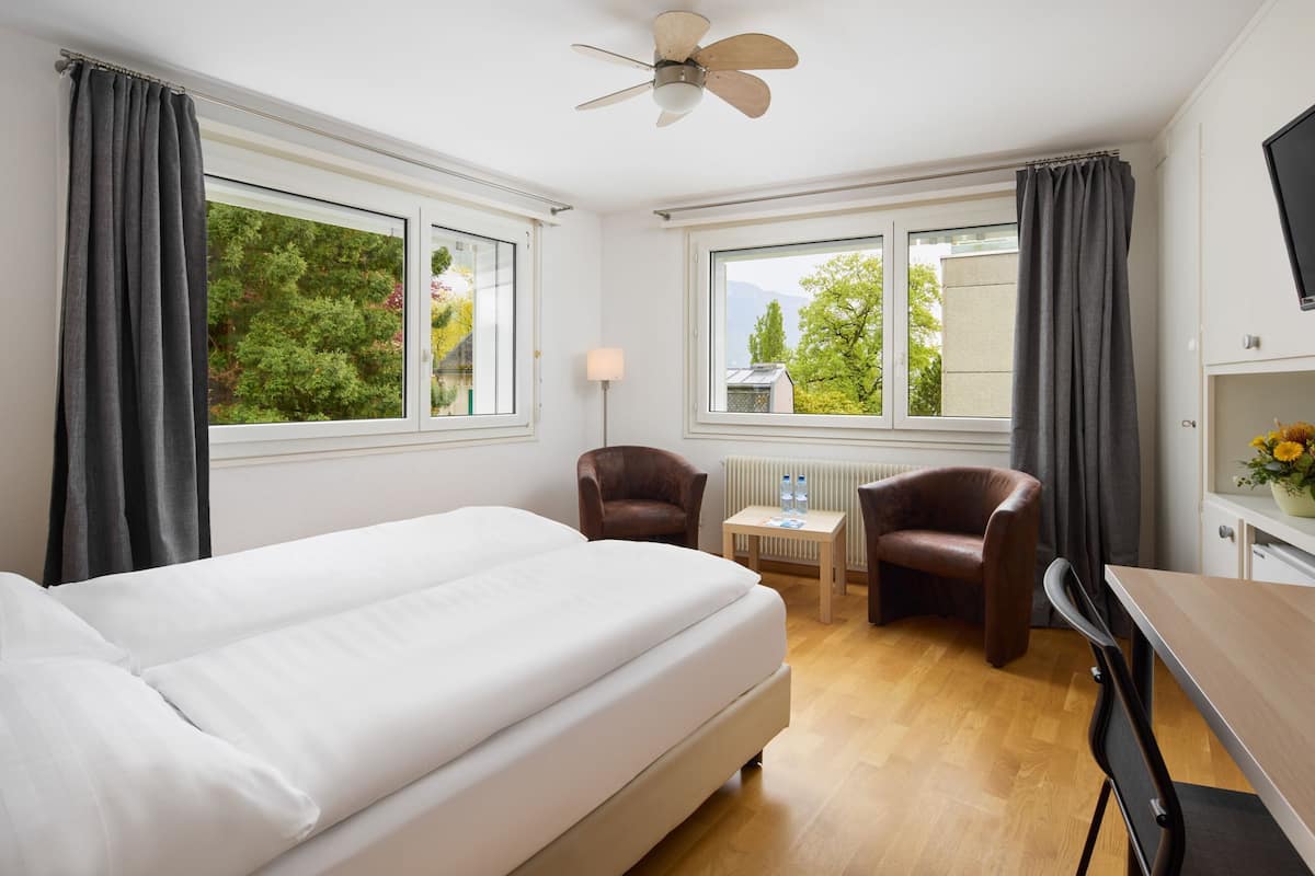 Quarto de hotel com cama, duas cadeiras, uma mesa, luminária e ventilador de teto.