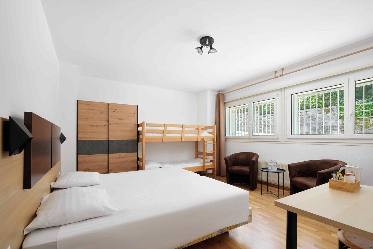 Quarto com uma cama beliche, uma escrivaninha, duas cadeiras, um radiador e uma janela com vista para o verde.