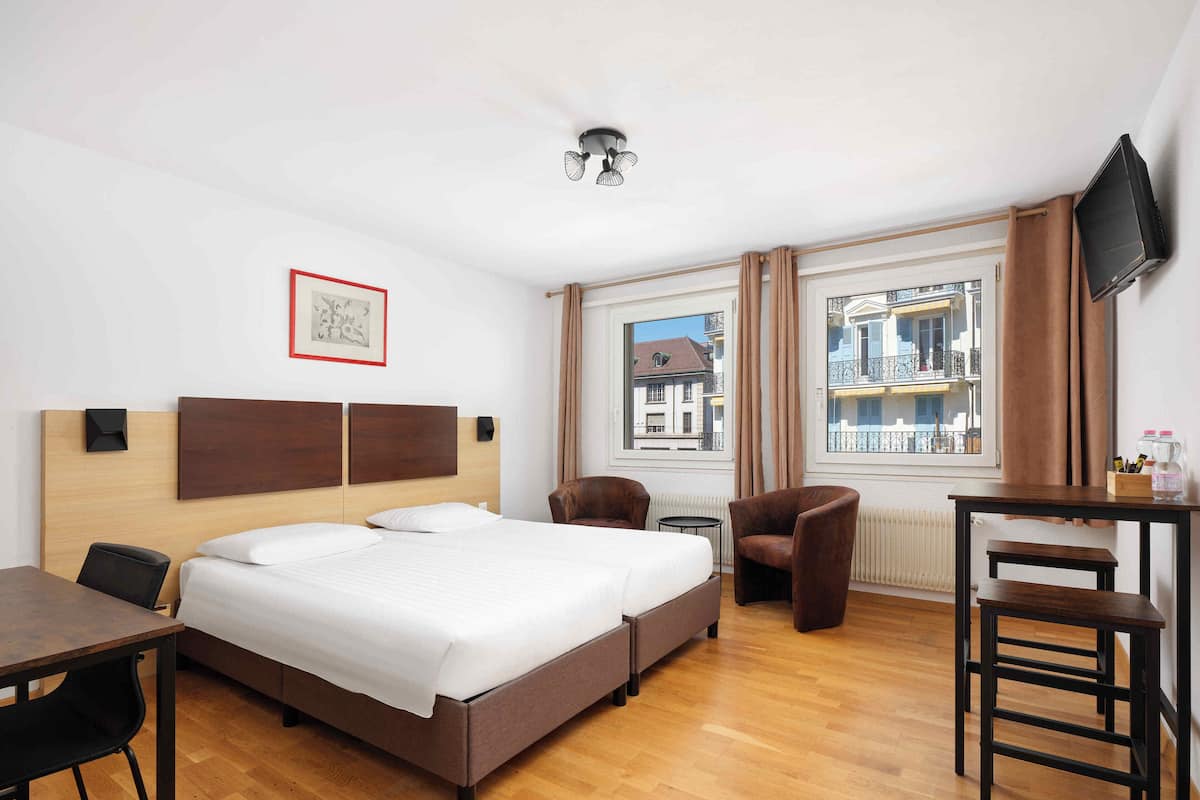 Quarto de hotel com uma cama grande, uma escrivaninha, duas cadeiras, uma televisão e uma janela com vista para edifícios.