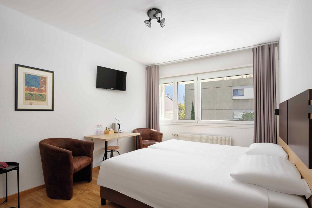 Quarto de hotel com cama, duas poltronas castanhas, escrivaninha com televisão de tela plana, quadro emoldurado e janela com cortinas.