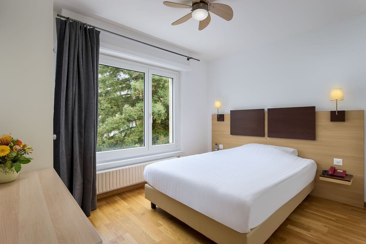 Quarto de hotel moderno com uma cama grande, um ventilador de teto, uma janela com cortinas, uma mesinha de cabeceira com abajur e uma pequena prateleira com um telefone e um vaso de flores.