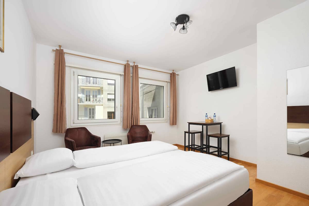Quarto de hotel com uma cama grande, duas cadeiras, uma mesinha, televisão e janela com cortinas.