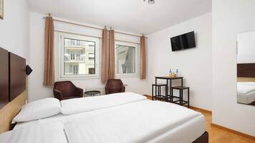 Quarto de hotel com uma cama grande, duas cadeiras, uma mesinha, televisão e janela com cortinas.