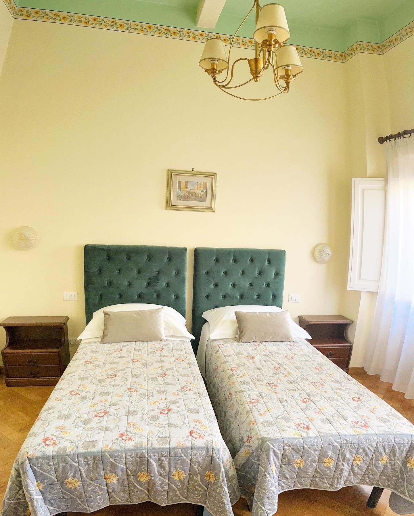 Quarto com duas camas, um lustre, um quadro emoldurado na parede e uma janela com cortinas.