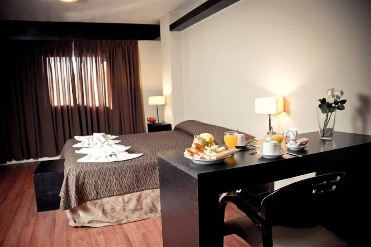 Quarto de hotel com uma cama, uma escrivaninha com itens de café da manhã, uma luminária e um vaso com flores.