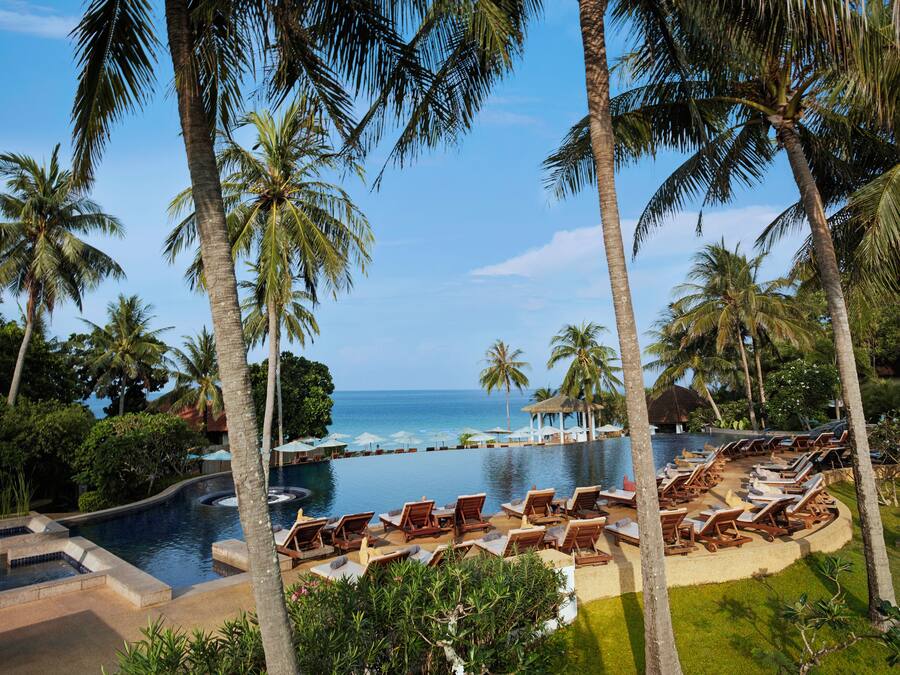 Uma área de piscina em resort, com espreguiçadeiras, palmeiras e vista para o oceano.