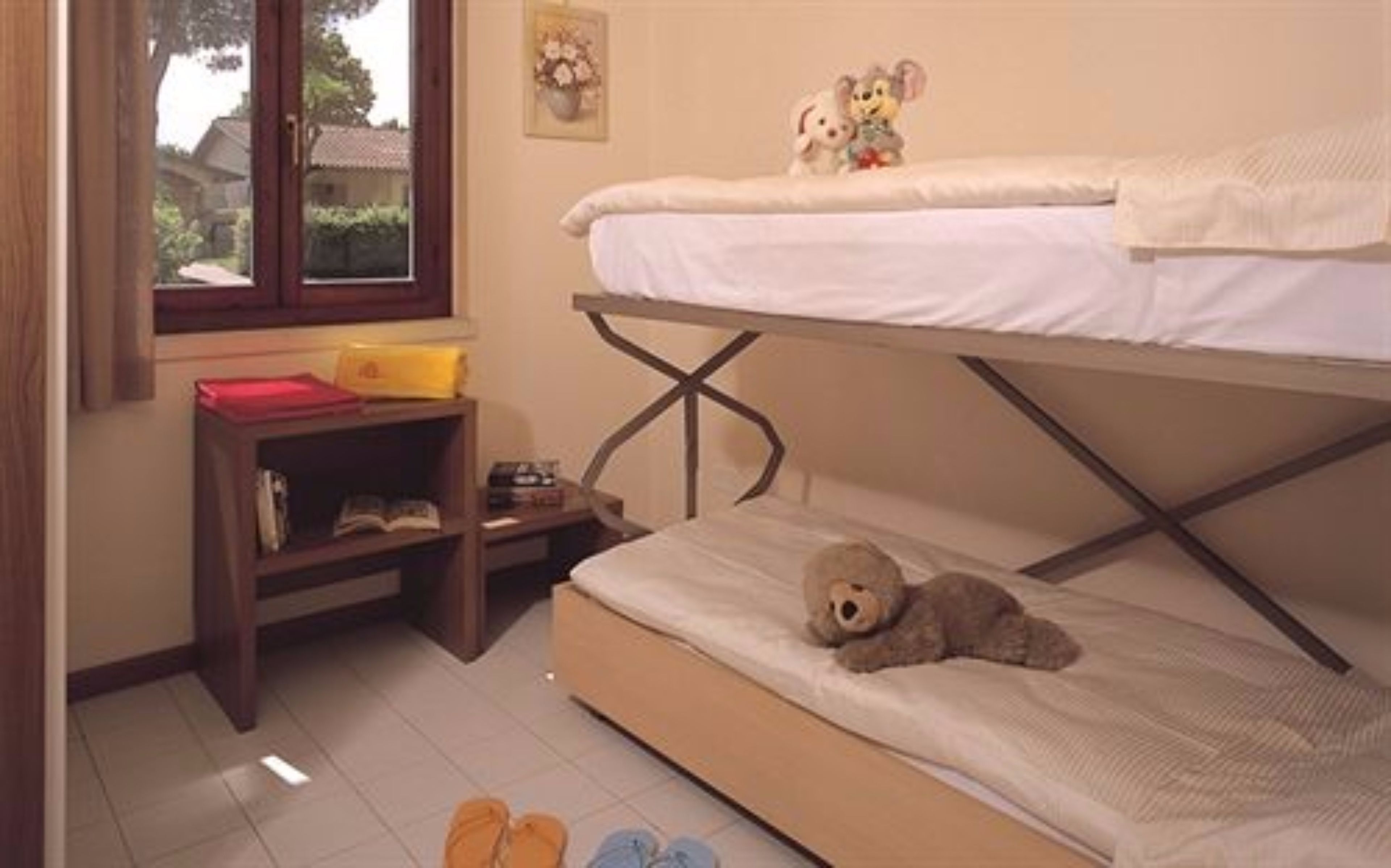 Um quarto de dormitório com uma cama de beliche, uma pequena escrivaninha com cadeira, um urso de pelúcia na cama inferior e uma janela com vista para uma casa.