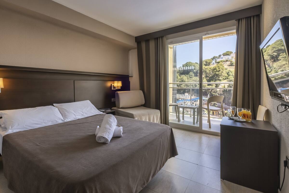 Quarto de hotel com uma cama grande, duas mesas de cabeceira, uma escrivaninha com cadeira e uma varanda com mesa e cadeiras.