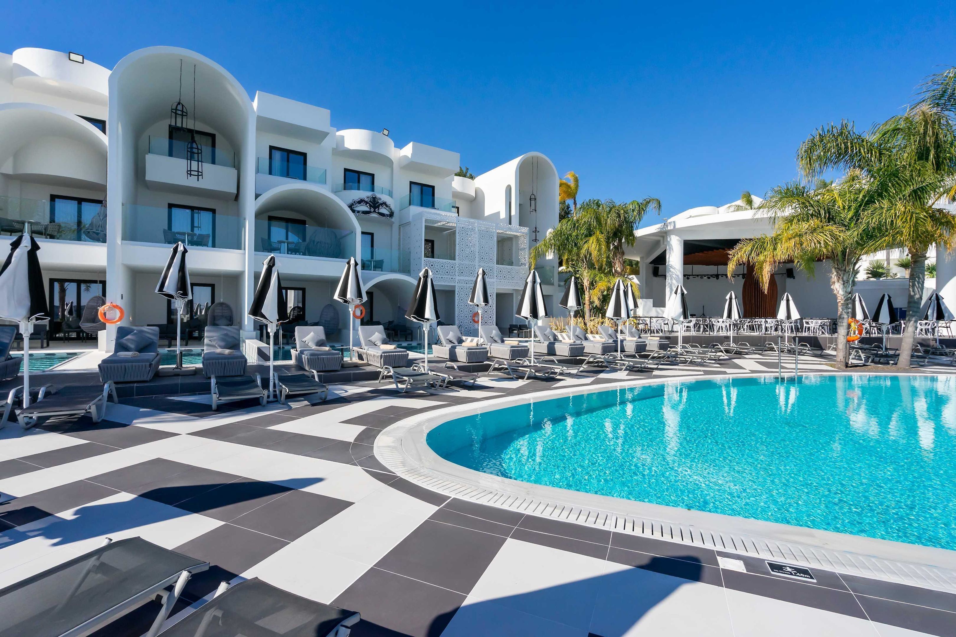 Uma área de piscina com espreguiçadeiras e guarda-sóis, rodeada por edifícios modernos com varandas.