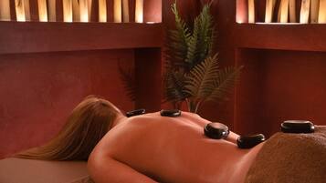 Uma pessoa recebendo uma massagem nas costas com pedras aquecidas, em um quarto com painéis de madeira e uma planta ao fundo.