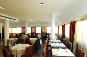 Uma ampla sala de jantar com várias mesas e cadeiras, janelas grandes com cortinas e um teto alto com iluminação embutida.