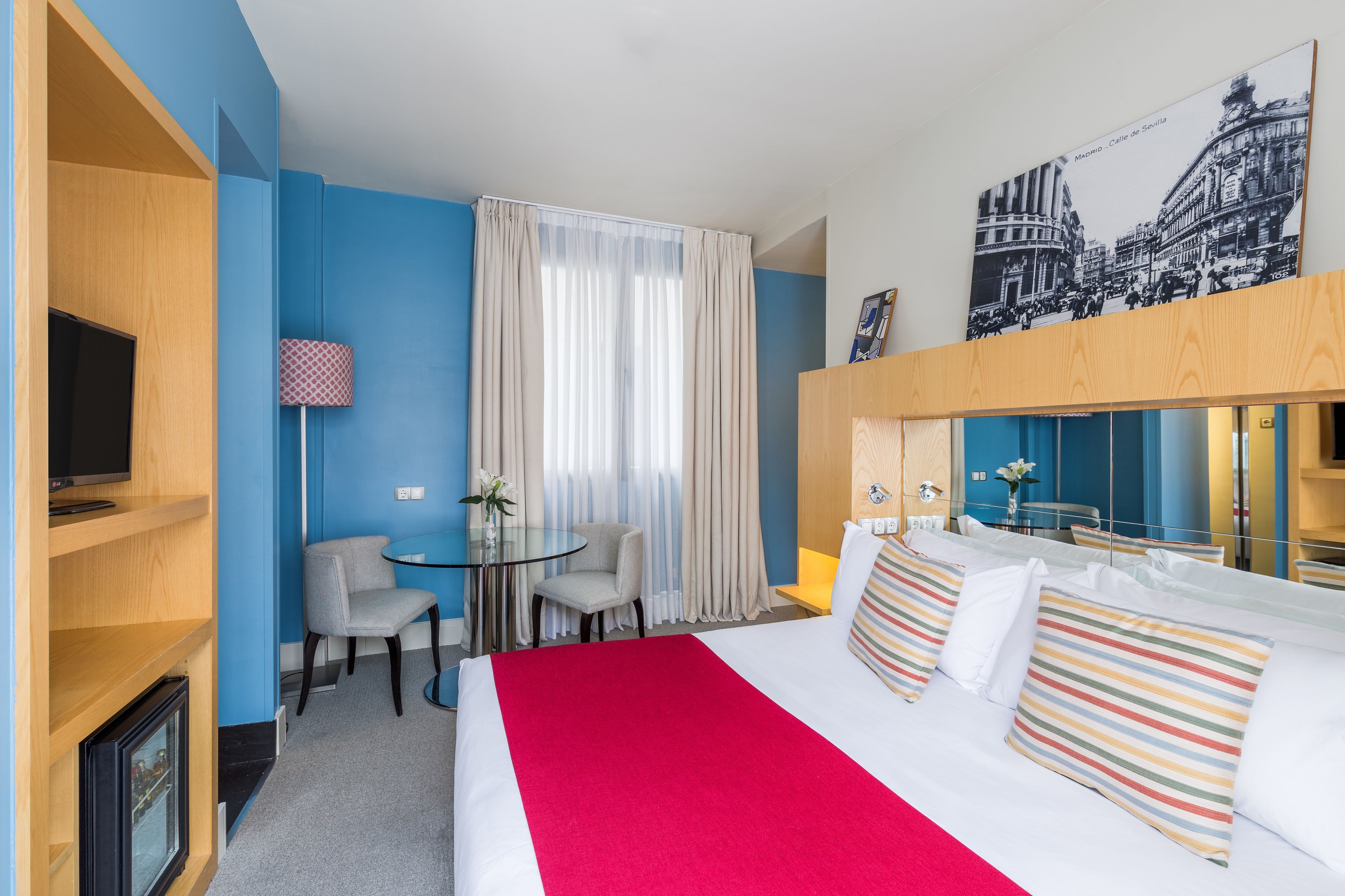 Quarto de hotel com cama, televisão, escrivaninha com cadeira e janela com cortinas.