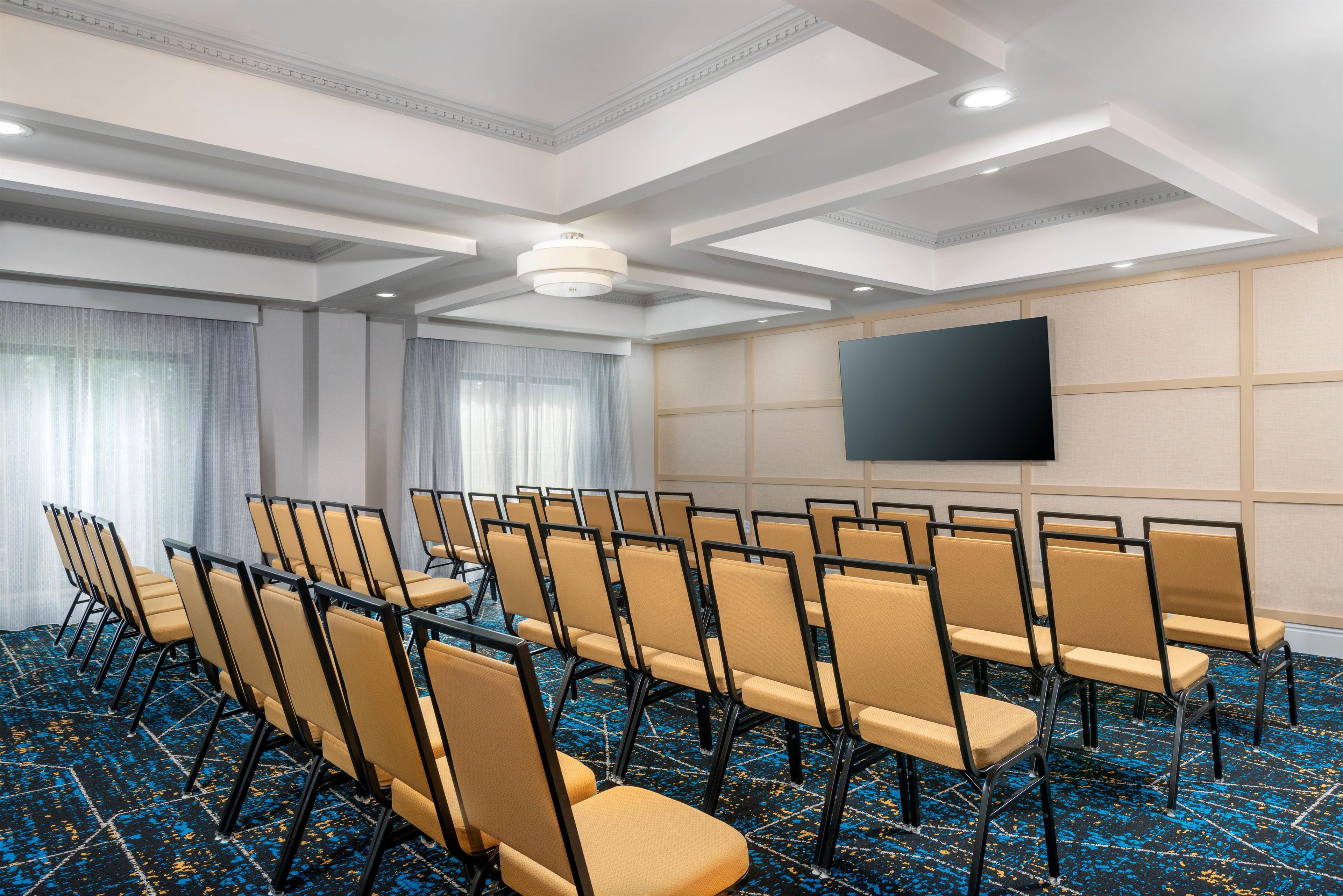 Uma sala de conferência com fileiras de cadeiras, uma tela grande e um carpete estampado.