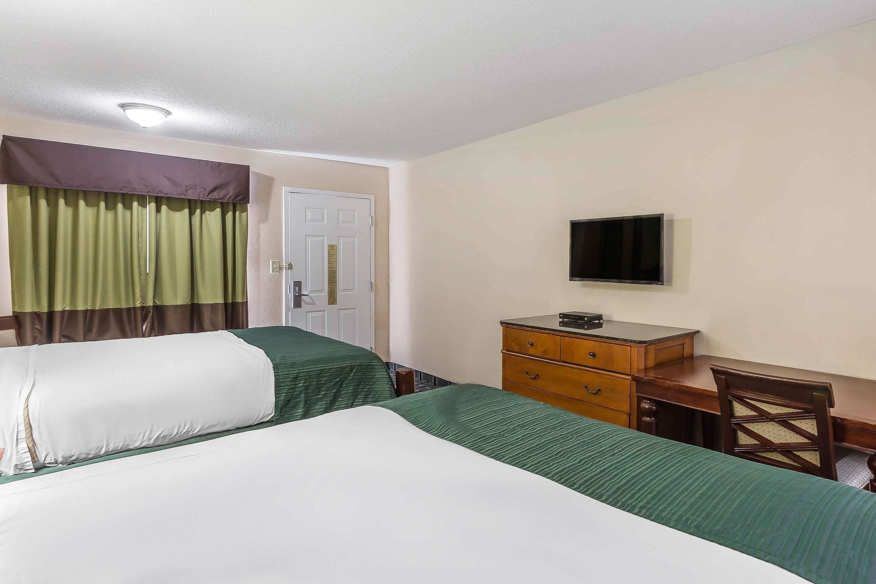 Quarto de hotel com duas camas, uma escrivaninha, uma cadeira, uma televisão e um guarda-roupa.