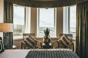 Quarto de hotel com uma cama grande, duas cadeiras, um vaso e vista para uma paisagem montanhosa através de três janelas.