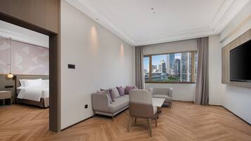 Quarto de hotel moderno com cama, sofá, poltrona, televisão e uma grande janela com vista para a cidade.