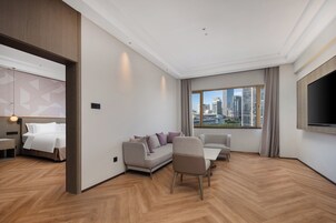 Quarto de hotel moderno com cama, sofá, poltrona, televisão e uma grande janela com vista para a cidade.