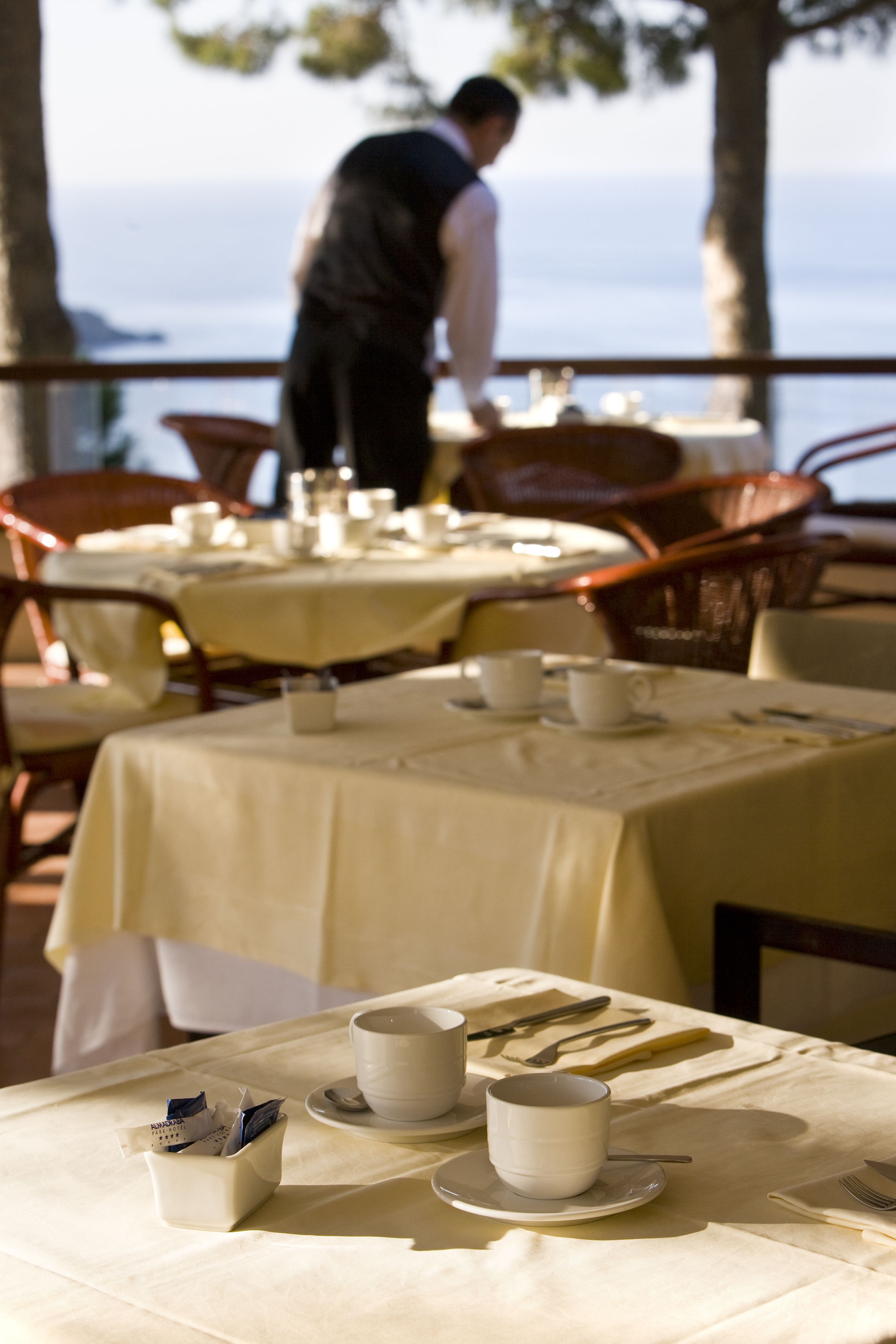 Uma sala de jantar com mesas preparadas para o serviço, um garçom ao fundo e vista para o mar.