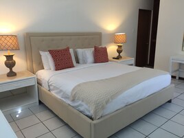 Uma cama bem arrumada com cabeceira bege, roupa de cama branca e duas almofadas vermelhas estampadas. Há duas mesas de cabeceira com abajures, uma de cada lado da cama.