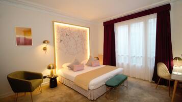 Quarto de hotel com uma cama grande, uma escrivaninha, uma cadeira, um banco e uma luminária fixa na parede.