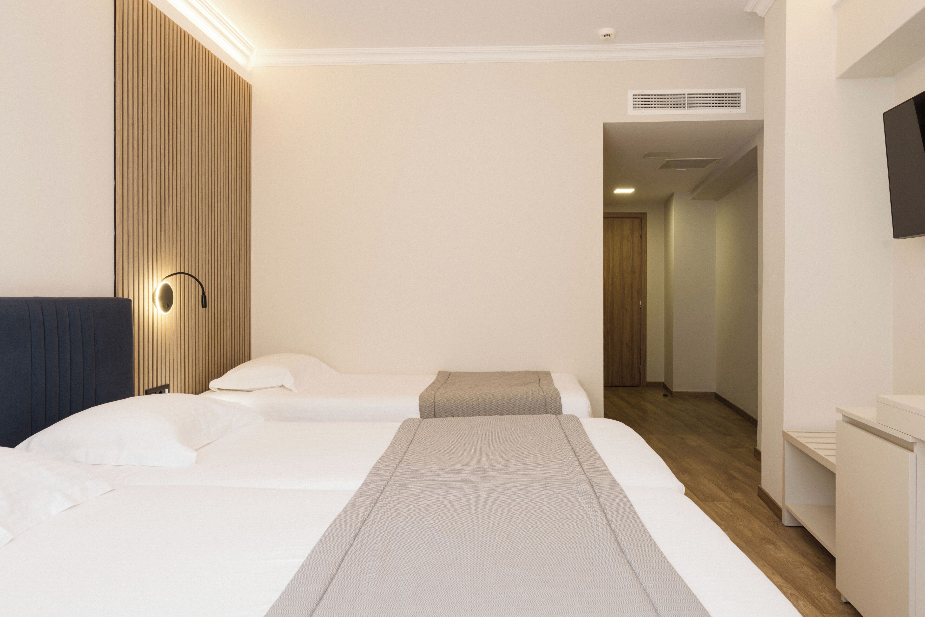 Quarto de hotel com duas camas, uma mesinha de cabeceira, uma luminária, uma televisão e parede de painel de madeira.